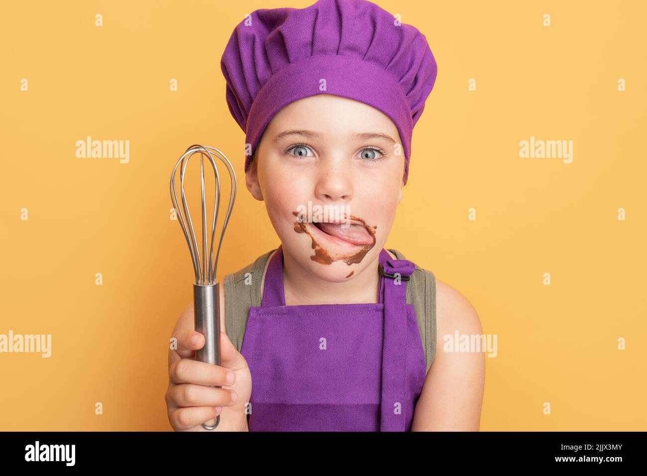 Cute smiling dirty boy in chef uniform looking away holding whisk while ...