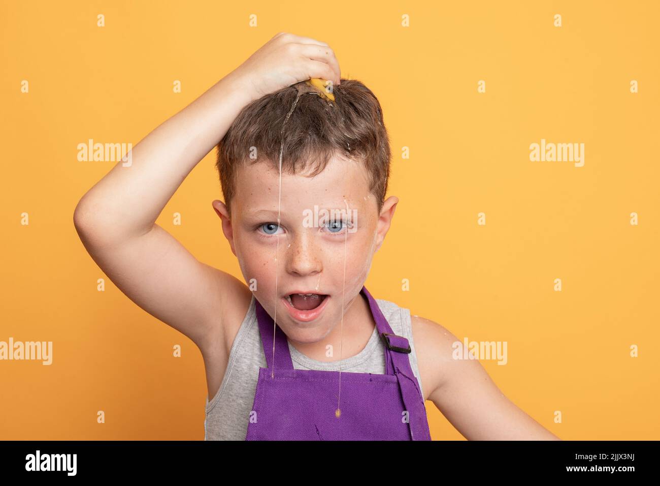 Happy boy in purple chef apron breaking raw egg on head while standing ...