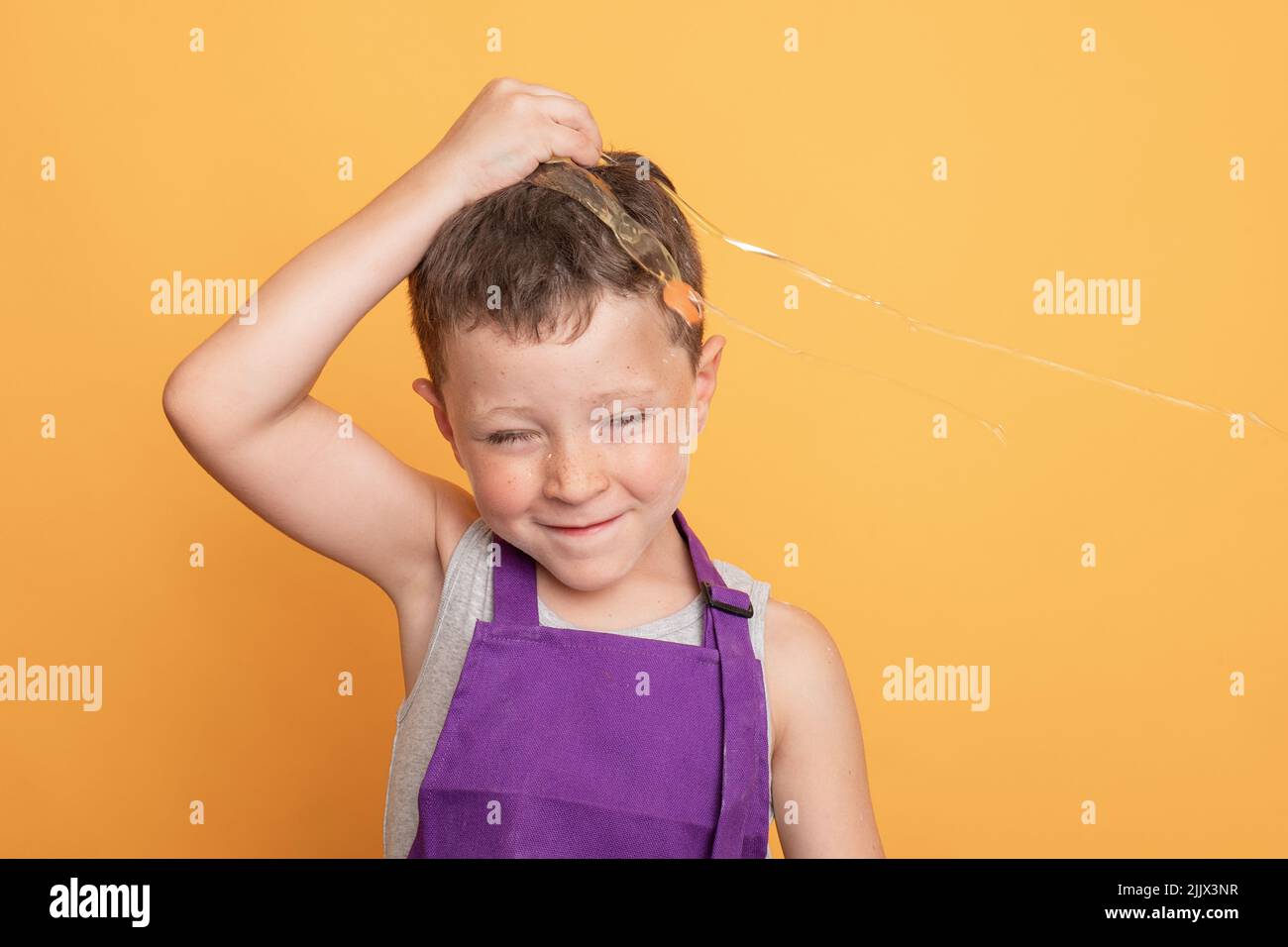 Happy boy in purple chef apron breaking raw egg on head while standing ...