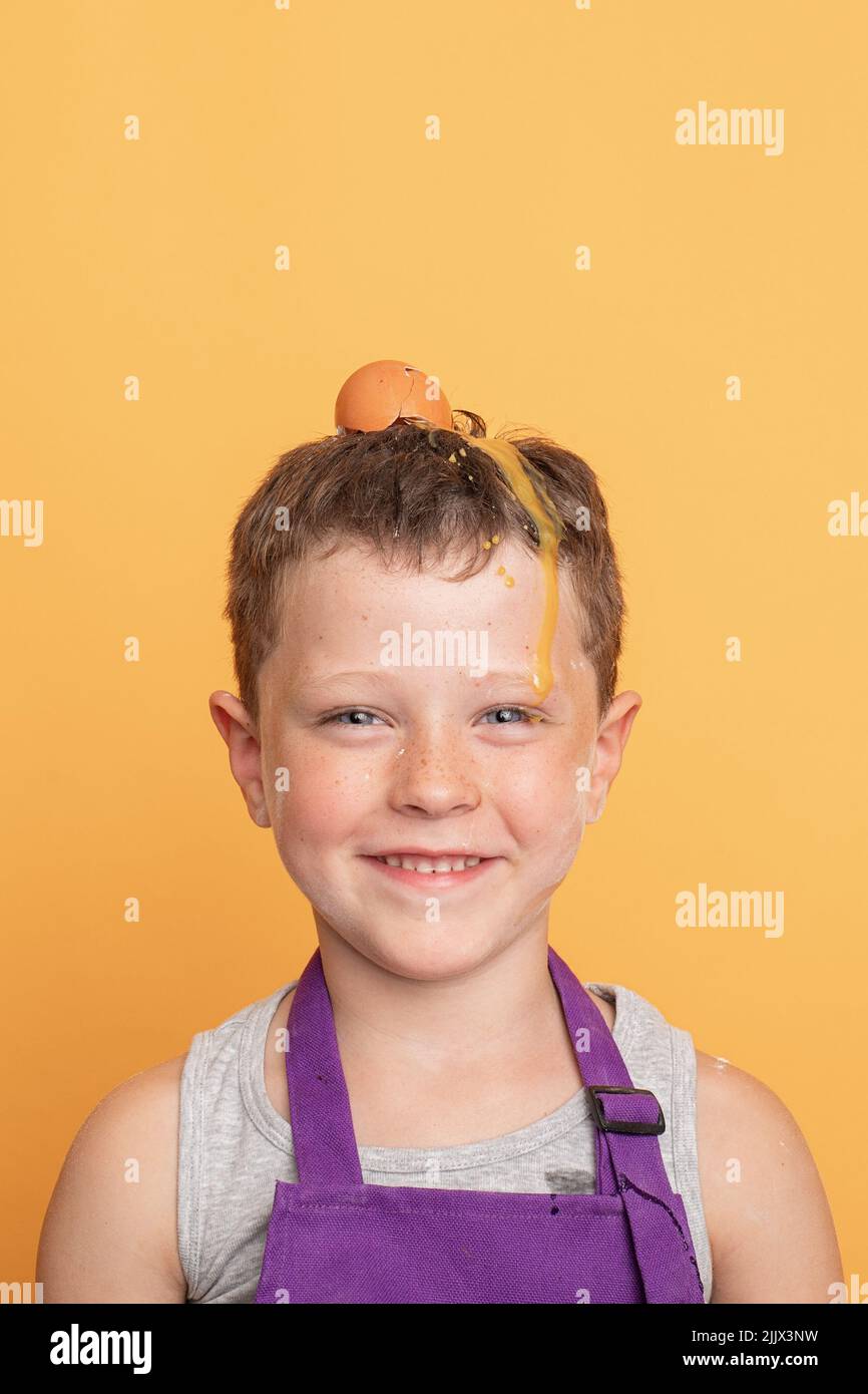 Funny boy in chef apron with broken raw egg on head looking at camera ...