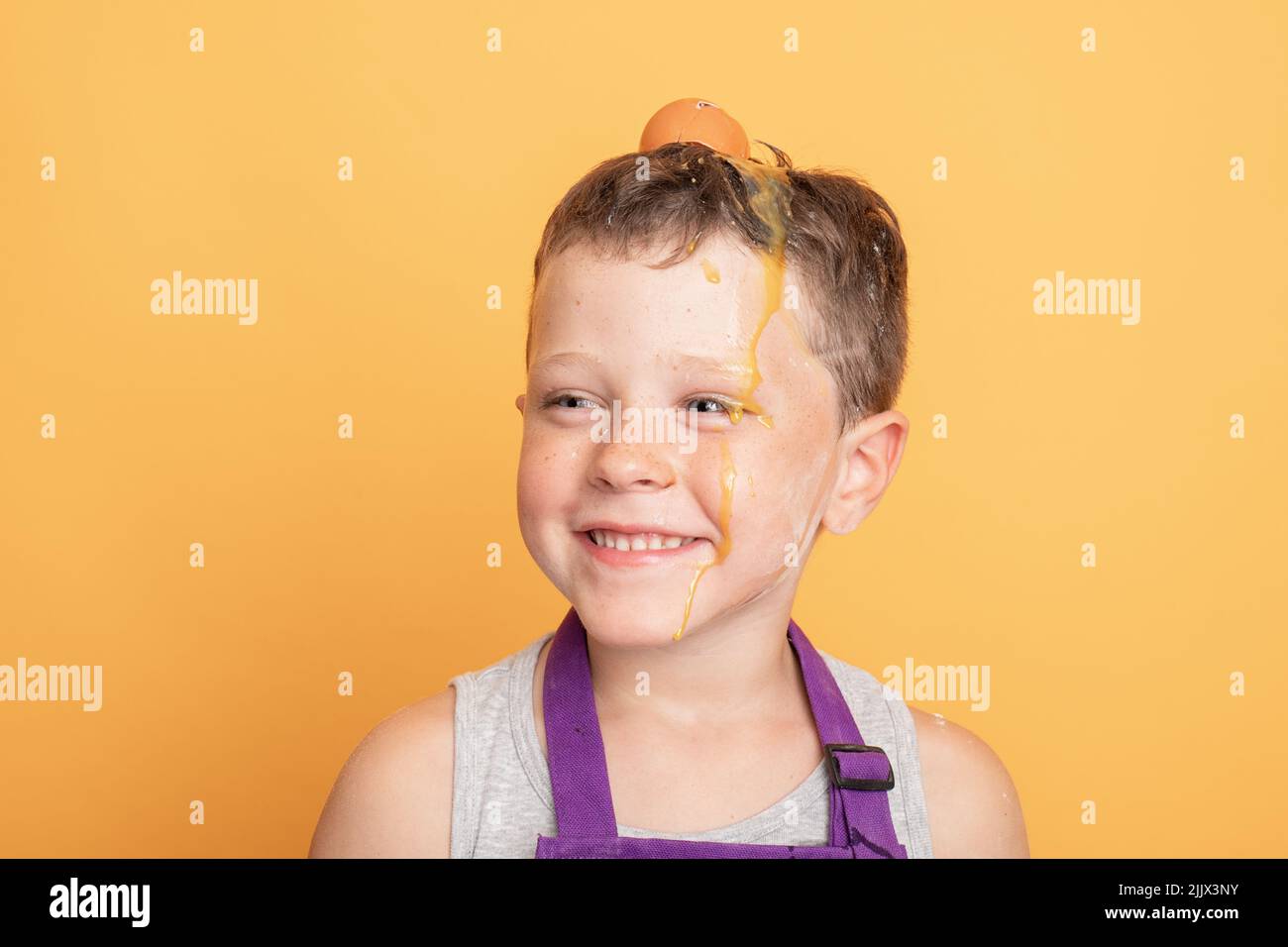 Funny boy in chef apron with broken raw egg on head looking away while ...