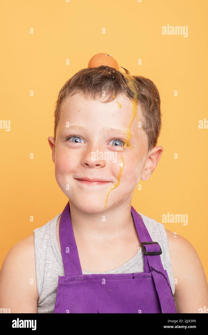 Funny boy in chef apron with broken raw egg on head looking at camera ...