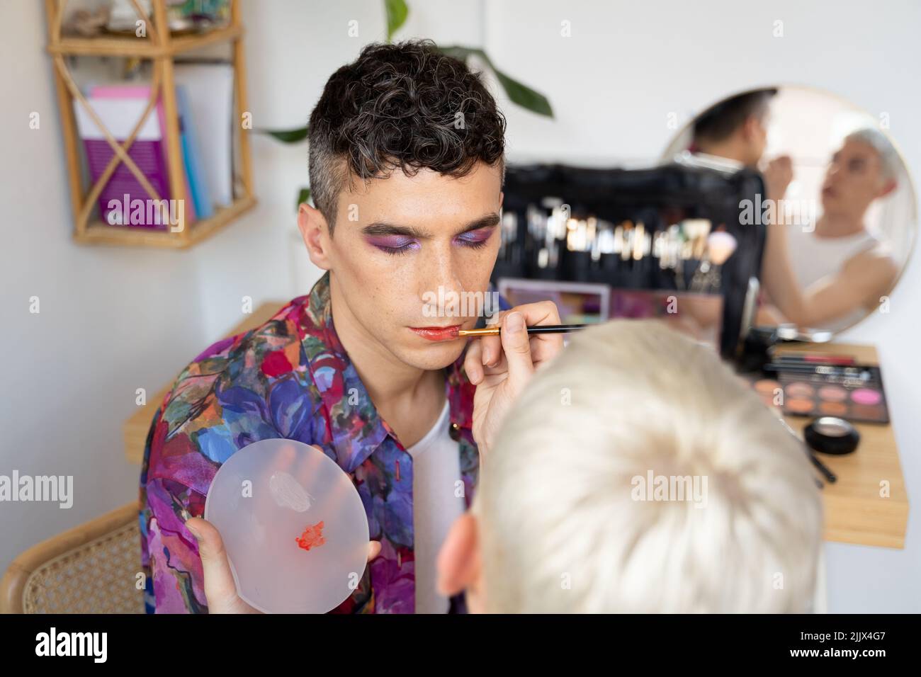 Young androgynous man getting makeup done by his boyfriend at dressing ...