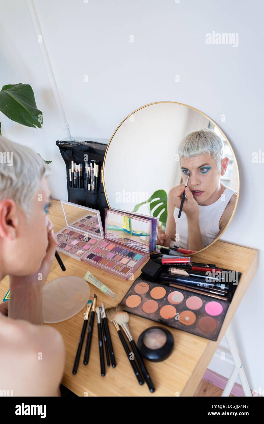 Transgender man wearing blue eyeshadow applying mascara while looking ...