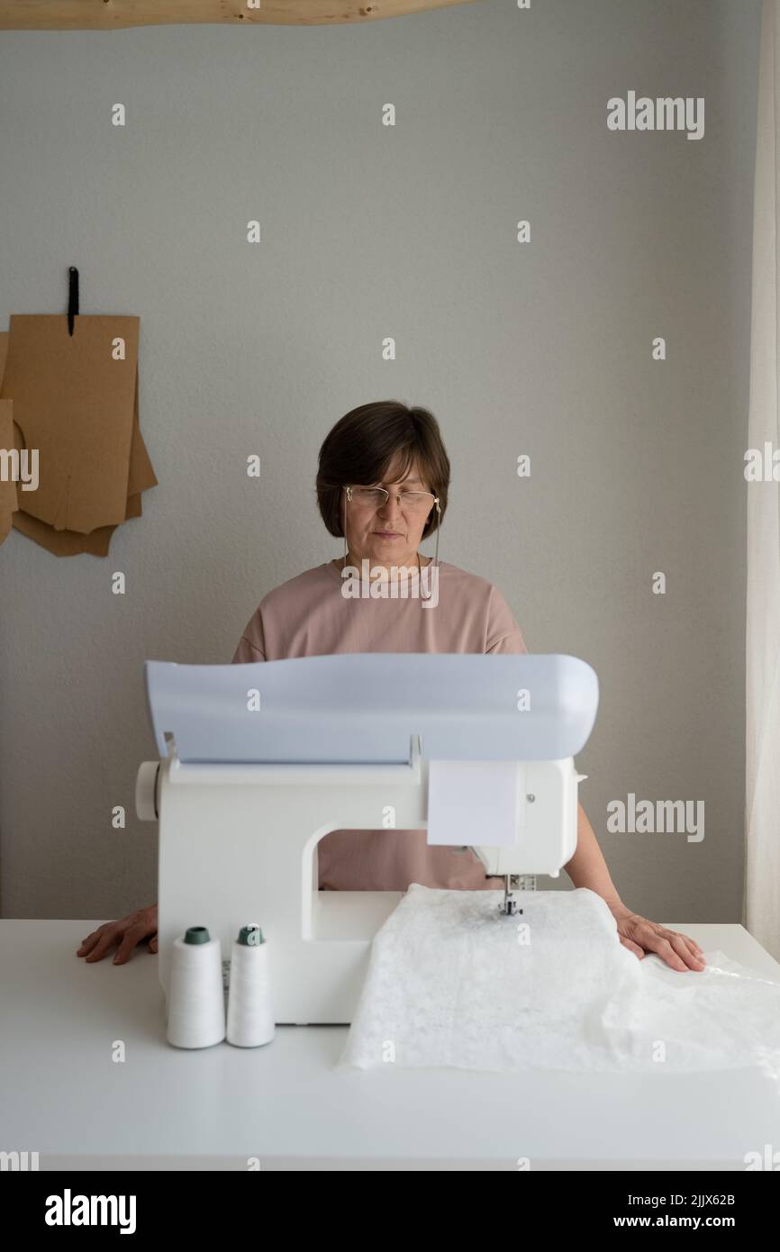 Concentrated female dressmaker in eyeglasses working on sewing machine ...