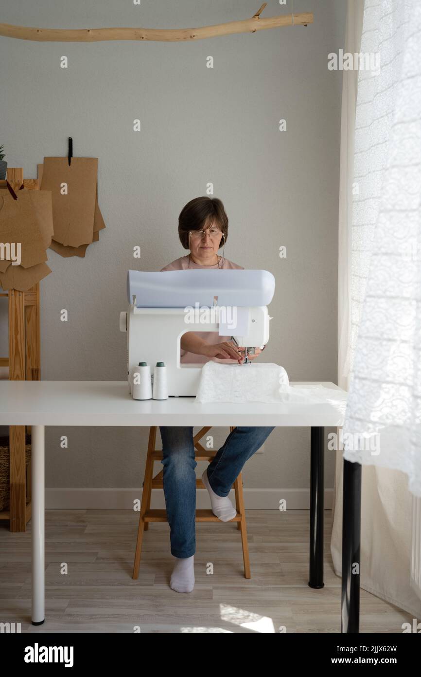 Concentrated female dressmaker in eyeglasses working on sewing machine ...