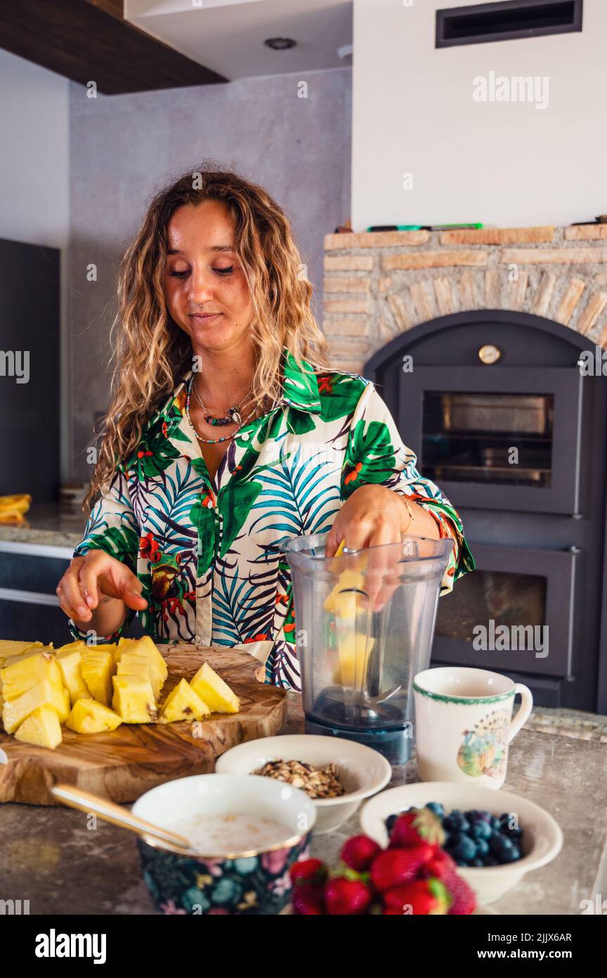 Focused female putting fresh chopped pineapple into blender bowl while ...