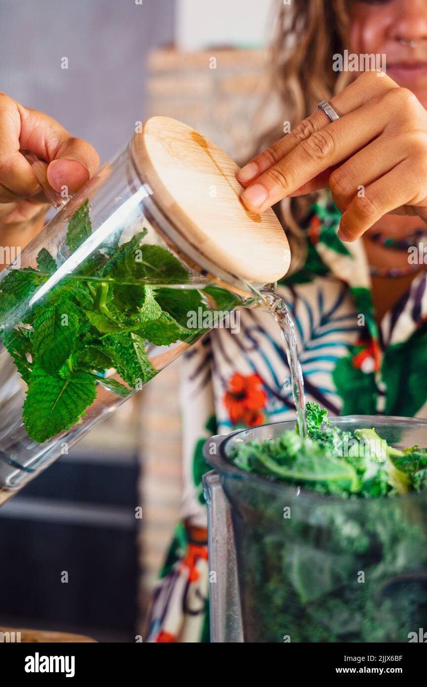 Crop unrecognizable female pouring refreshing water with mint into blender bowl with green kale ...