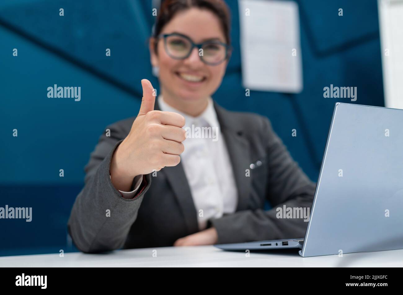 Smiling woman in a business suit shows thumb up while sitting at a desk ...