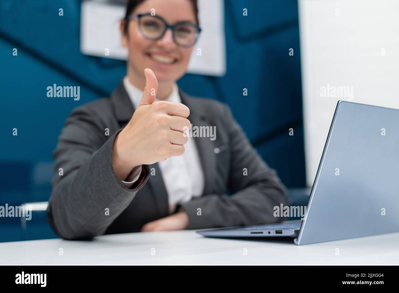 Smiling woman in a business suit shows thumb up while sitting at a desk ...