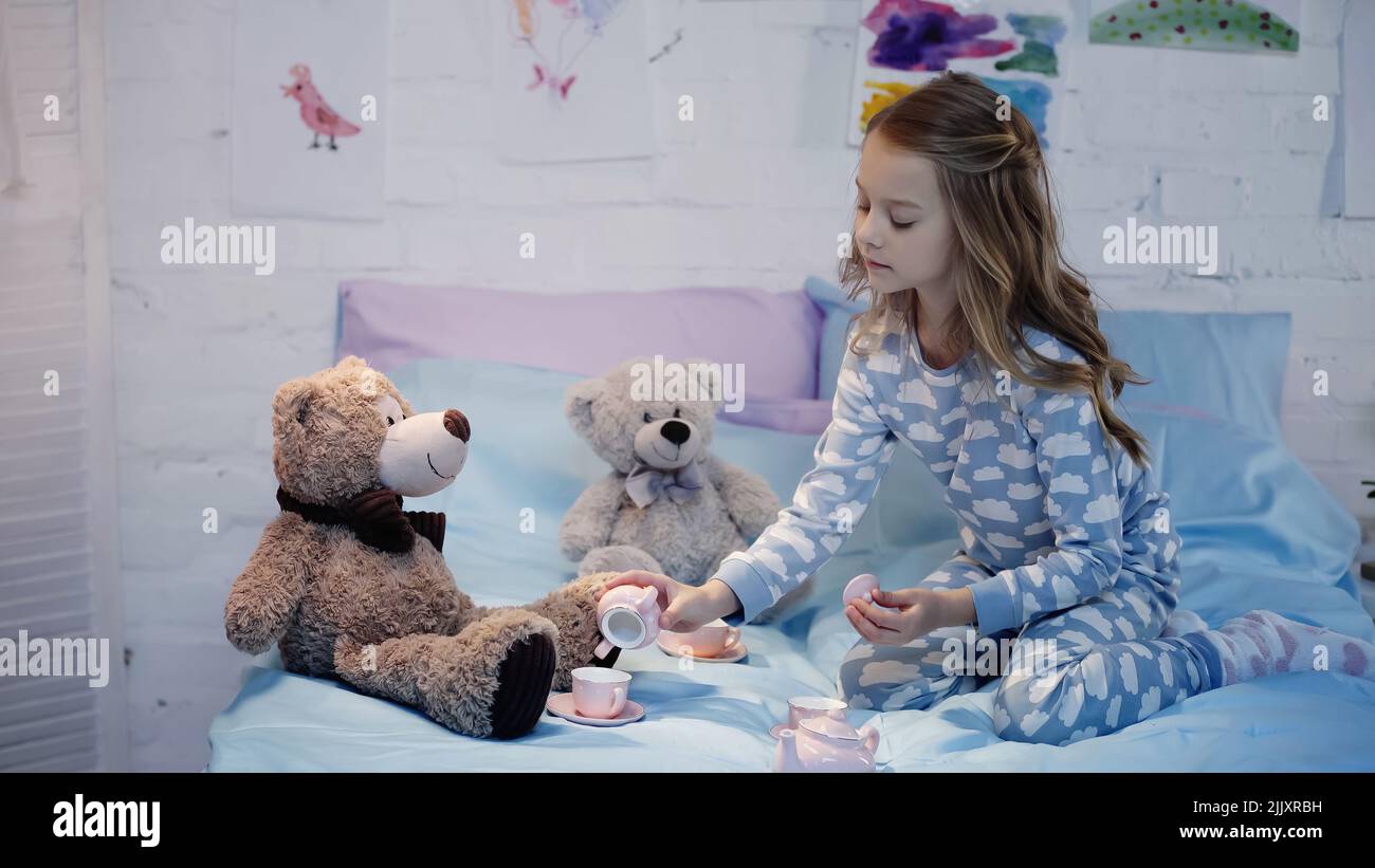 Child in pajama playing with tea cups and teddy bears on bed in evening ...