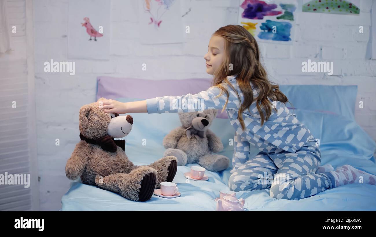 Side view of preteen kid touching soft toy near tea cups on bed in ...