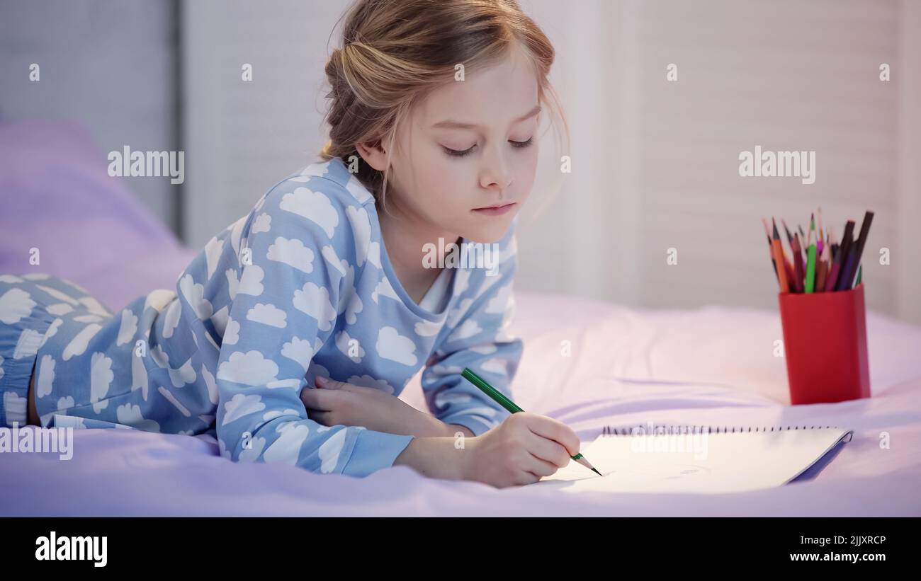 Preteen child in pajama drawing with color pencil on sketchbook on bed ...