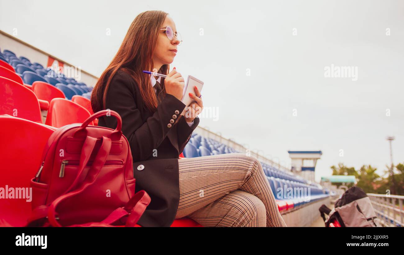 Young woman in glasses with notepad, pen sitting on stadium bleachers ...