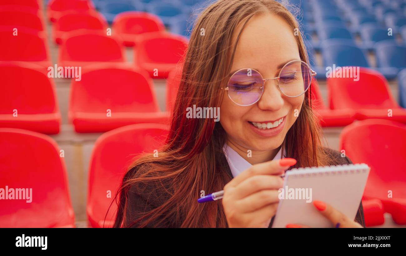 Young woman in glasses with notepad, pen sitting on stadium bleachers ...