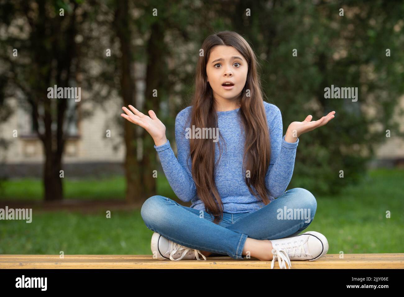 Shocked teenage girl shrugging shoulders sitting legs crossed on bench ...