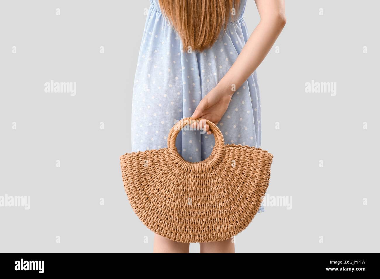 Young woman holding rattan bag behind her back on light background ...