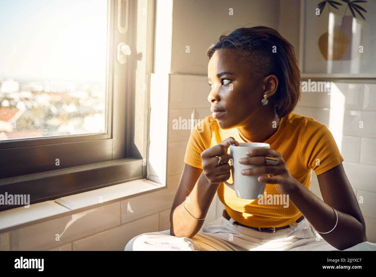 A thinking and coffee drinking black woman enjoys her morning routine while gazing outside a ...