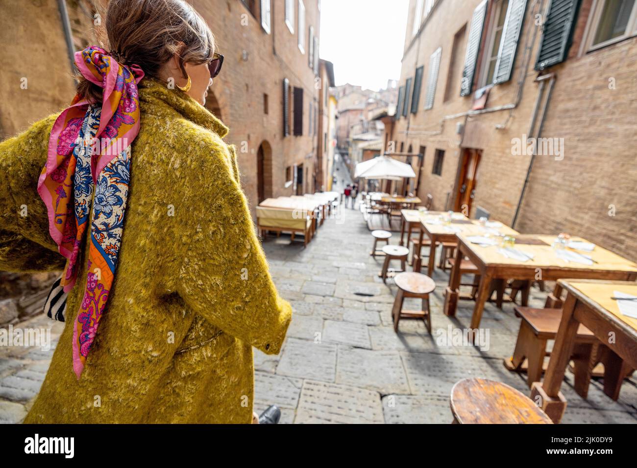 Sylish woman walks on narrow and cozy street in old town of Siena city ...