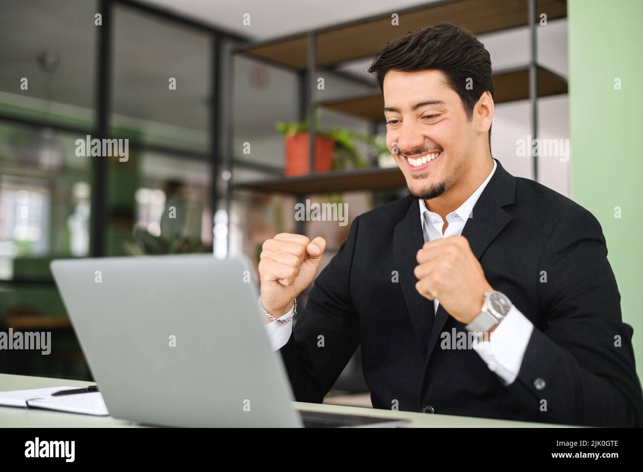 Excited Lucky Hispanic Young Businessman Sitting At The Desk In Front