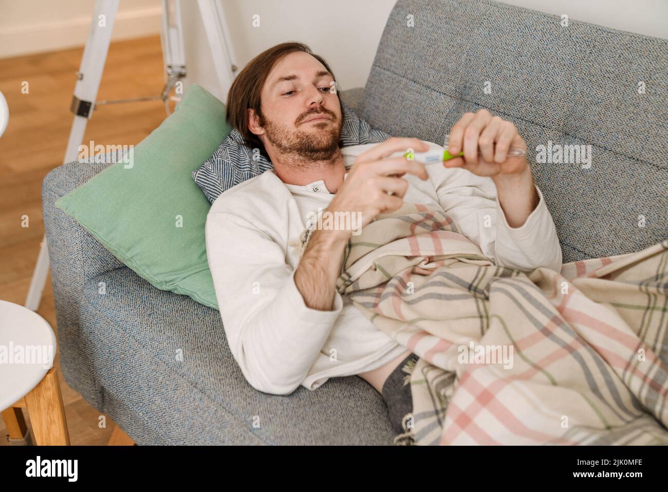 Ginger man feeling sick and measuring temperature while lying on couch ...