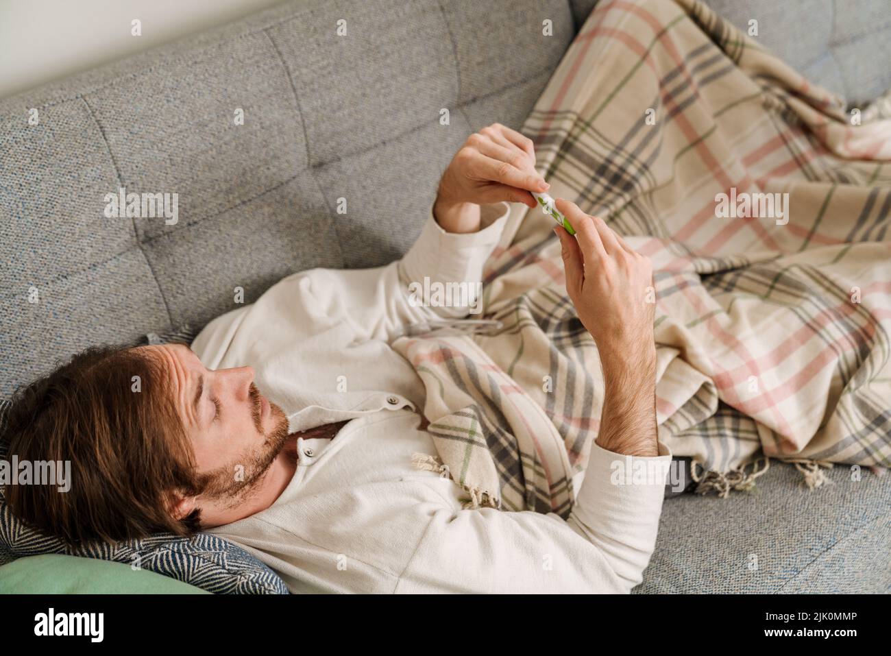 Ginger man feeling sick and measuring temperature while lying on couch ...