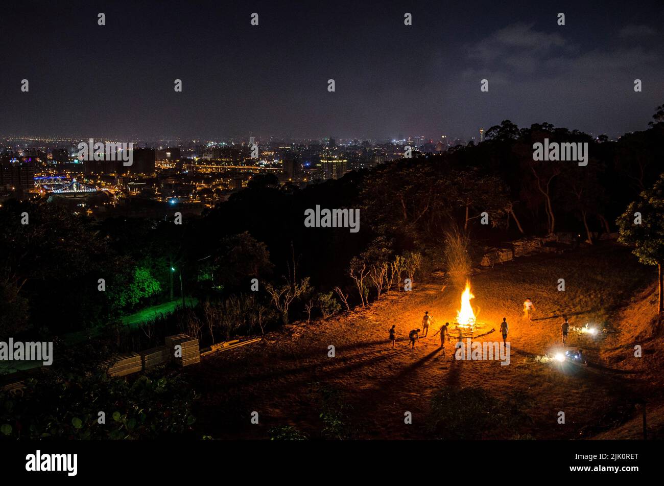 An aerial view of people around a bonfire on a hill against a city ...