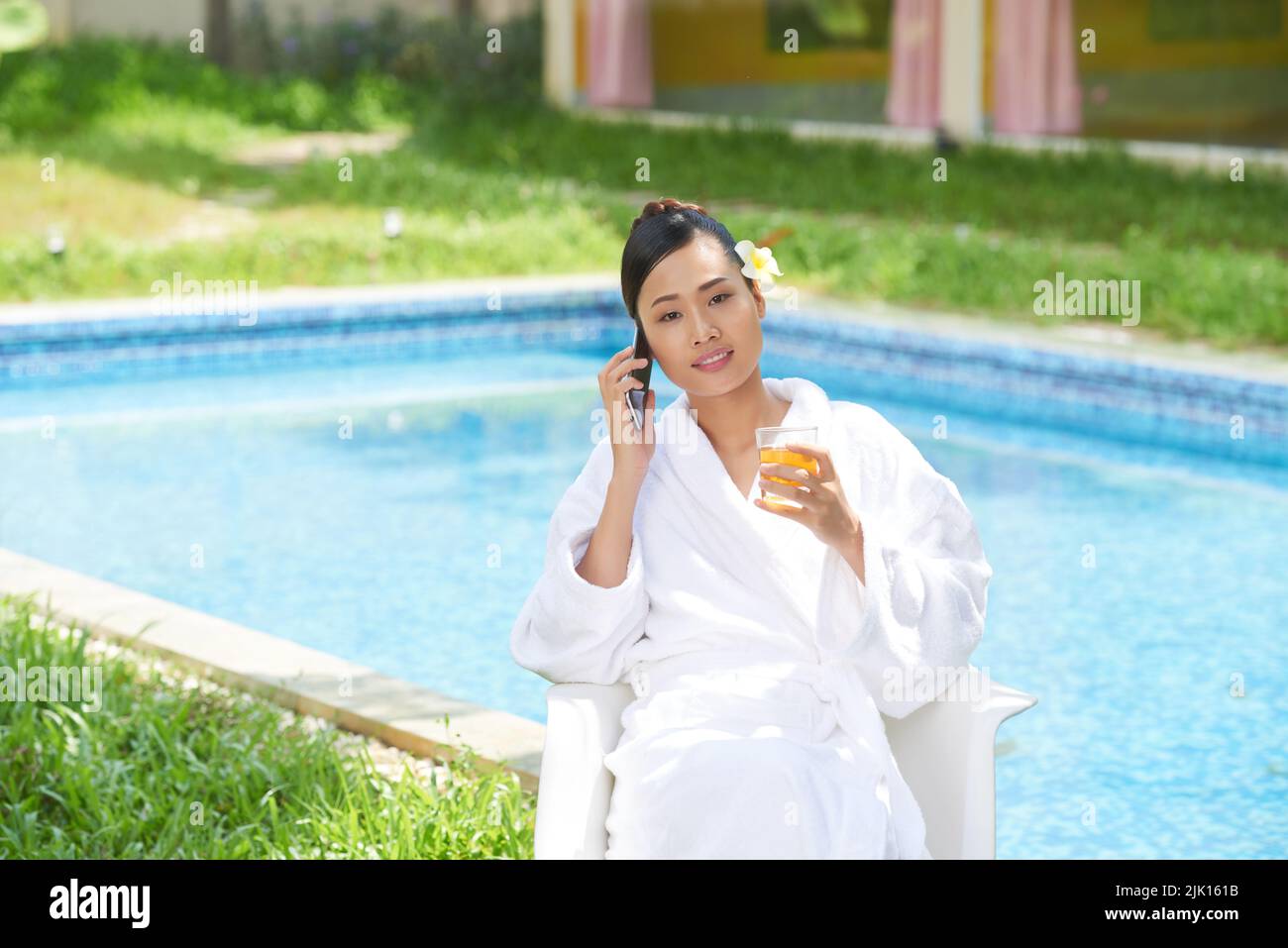 Lovely Vietnamese young woman sitting by the pool, drinking juice and ...