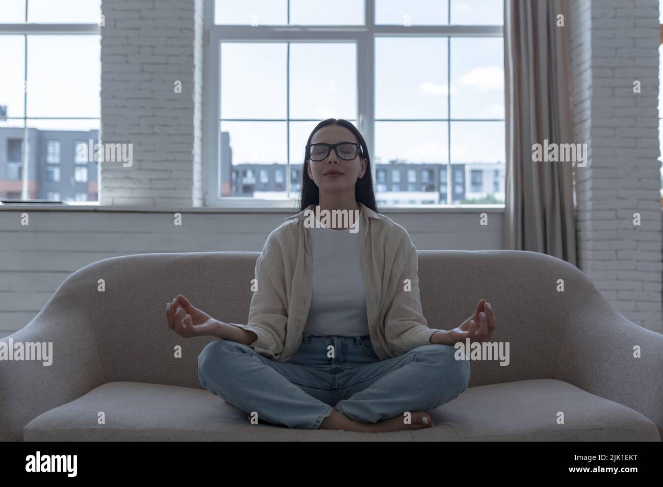 Woman alone at home meditating in front of window sitting on sofa ...