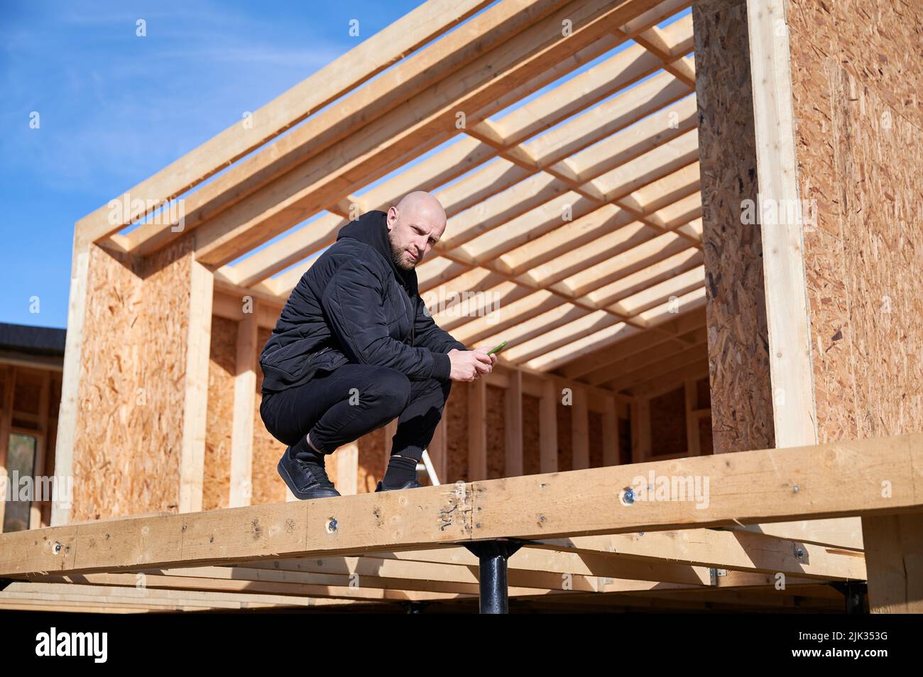 Male engineer building wooden frame house on pile foundation. Bald man standing on construction ...