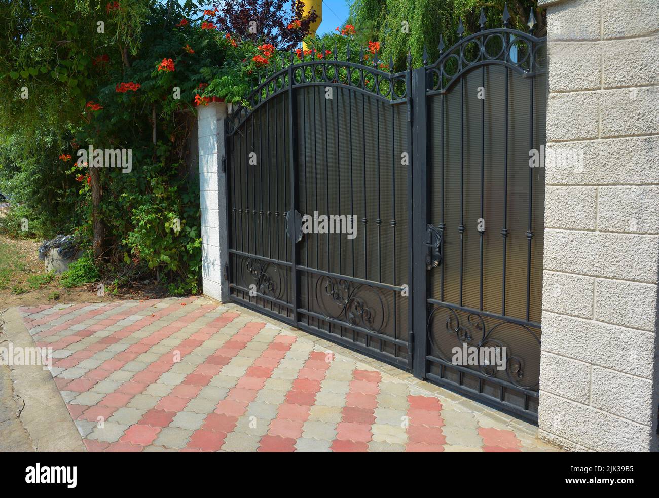 A metal wrought iron gate with brick fence posts, a paved driveway, and ...
