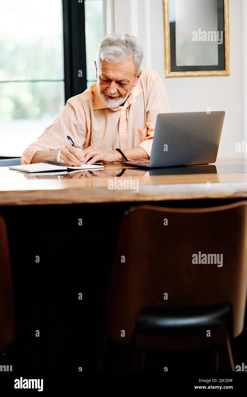 A happy senior student is sitting at home and writing down notes in a ...