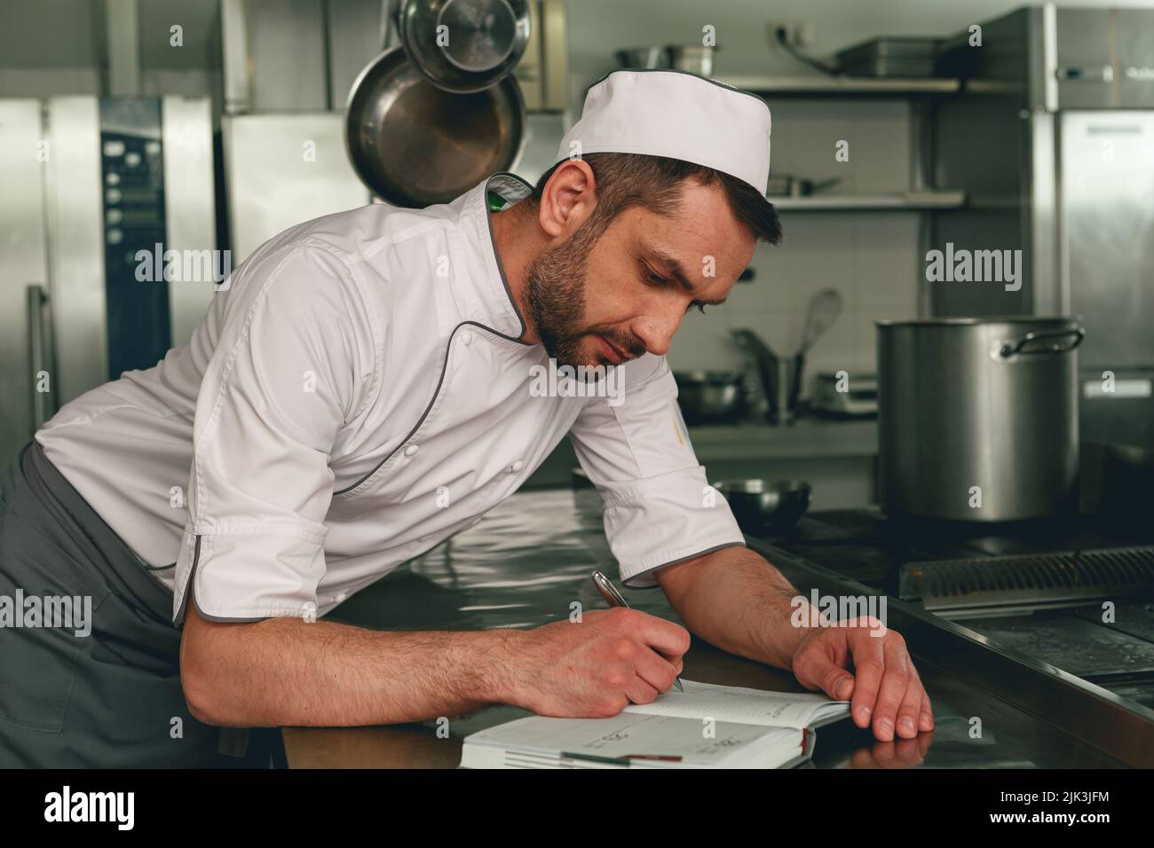 Smiling chef in uniform making notes in notebook standing on kitchen ...
