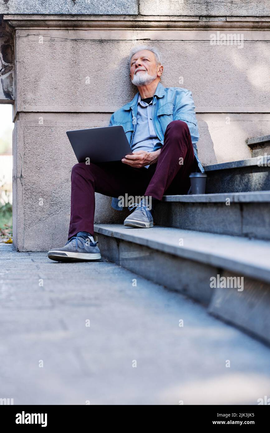 A senior student sits on the stairs of the university building and uses ...