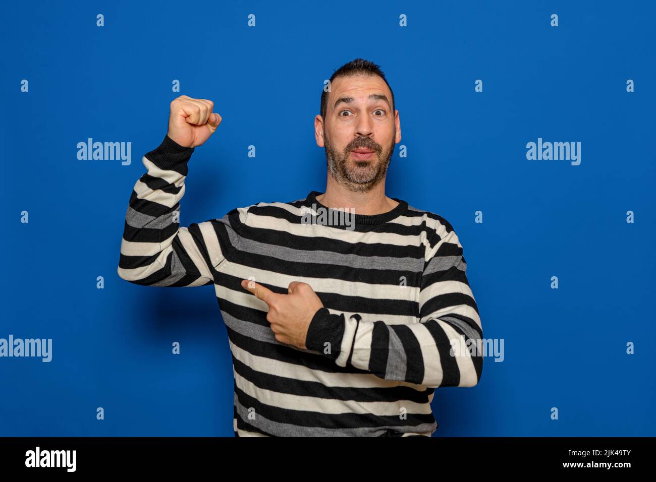Unshaven 40 year old man in striped pullover pointing finger at biceps ...