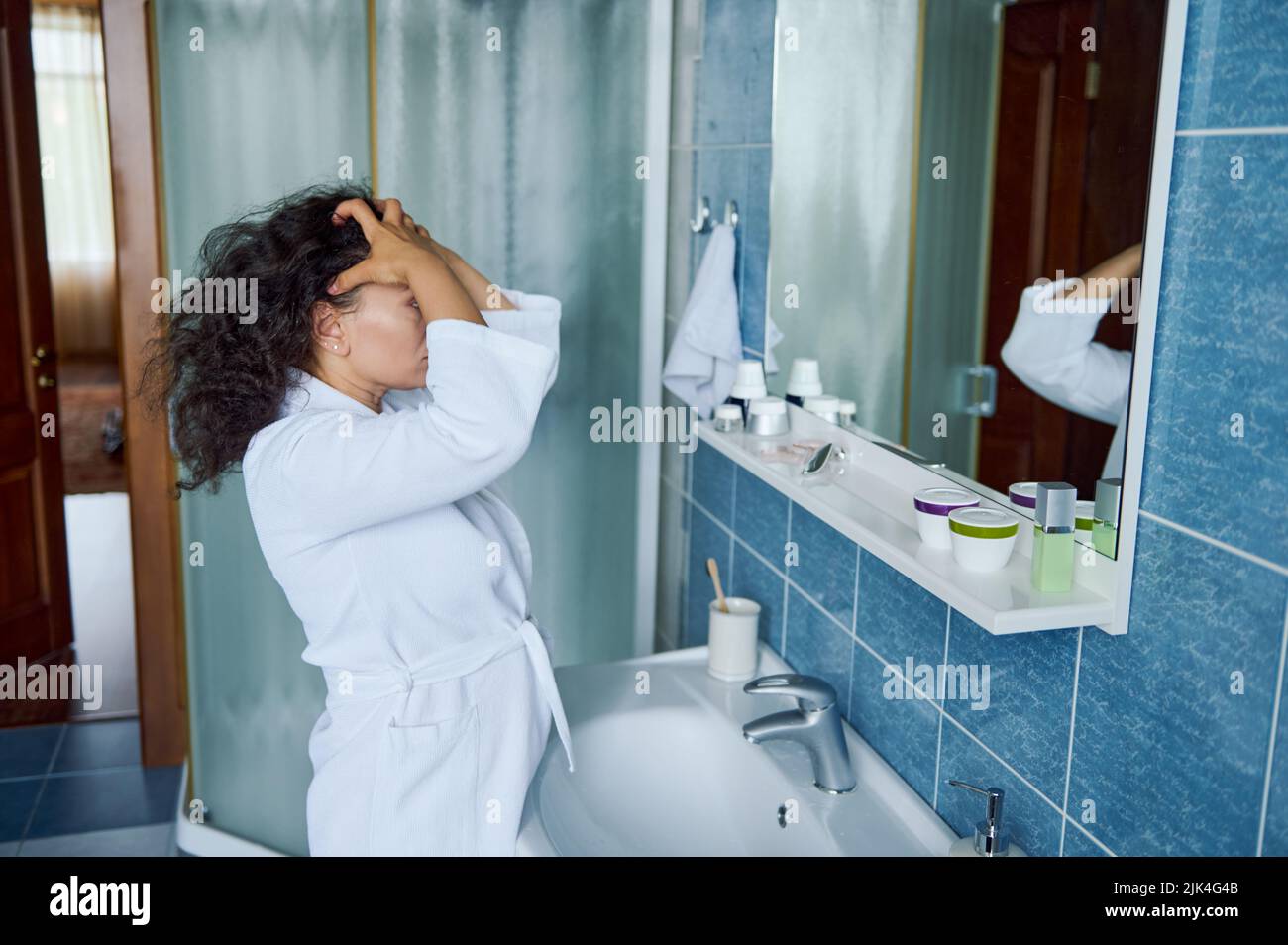 Cute woman in white bathrobe standing in bathroom reflected in mirror ...