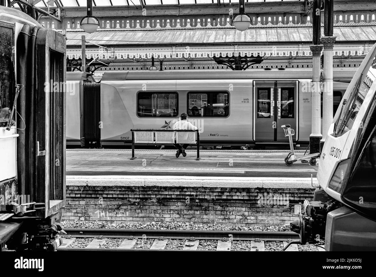 Seen through two trains, a man sits on a bench with a carriage in front ...