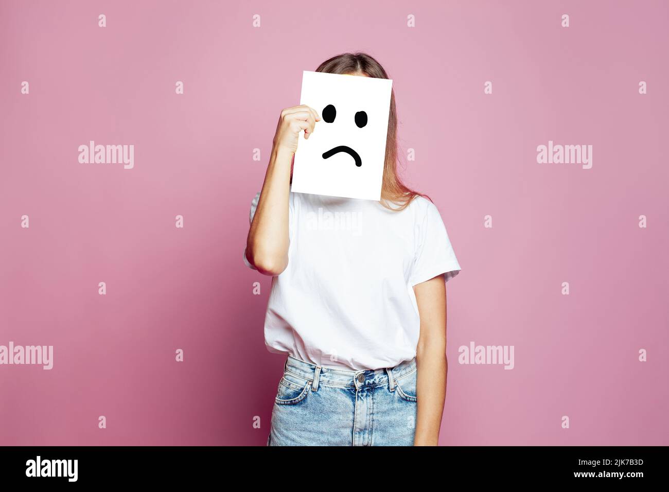 Young woman hiding her face using white paper with sad sign on pink ...