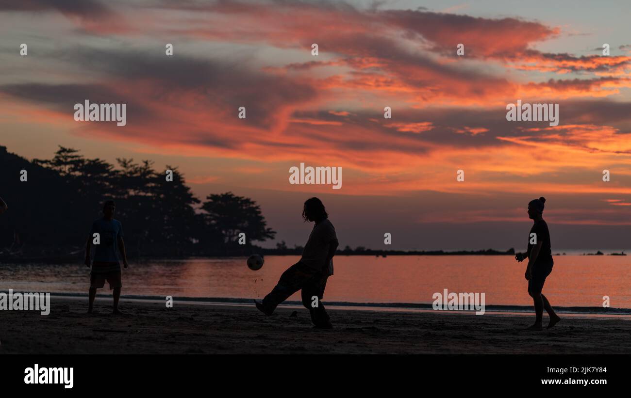 Sunset beach soccer. People in silhouette play football on the beach as ...