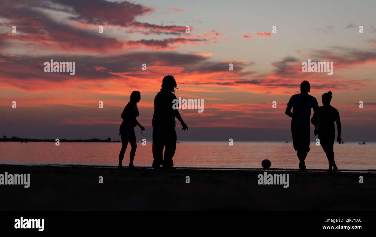 Sunset beach soccer. People in silhouette play football on the beach as ...