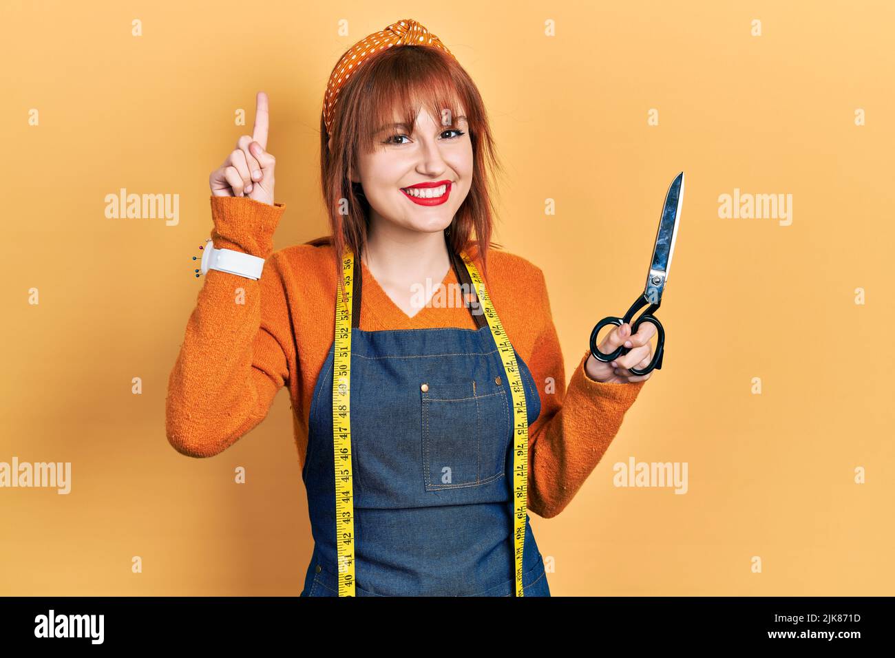 Redhead young woman dressmaker designer wearing atelier apron holding scissors smiling with an ...