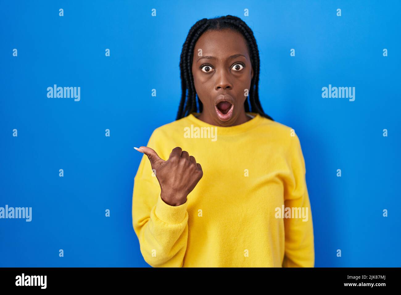 Beautiful black woman standing over blue background surprised pointing ...