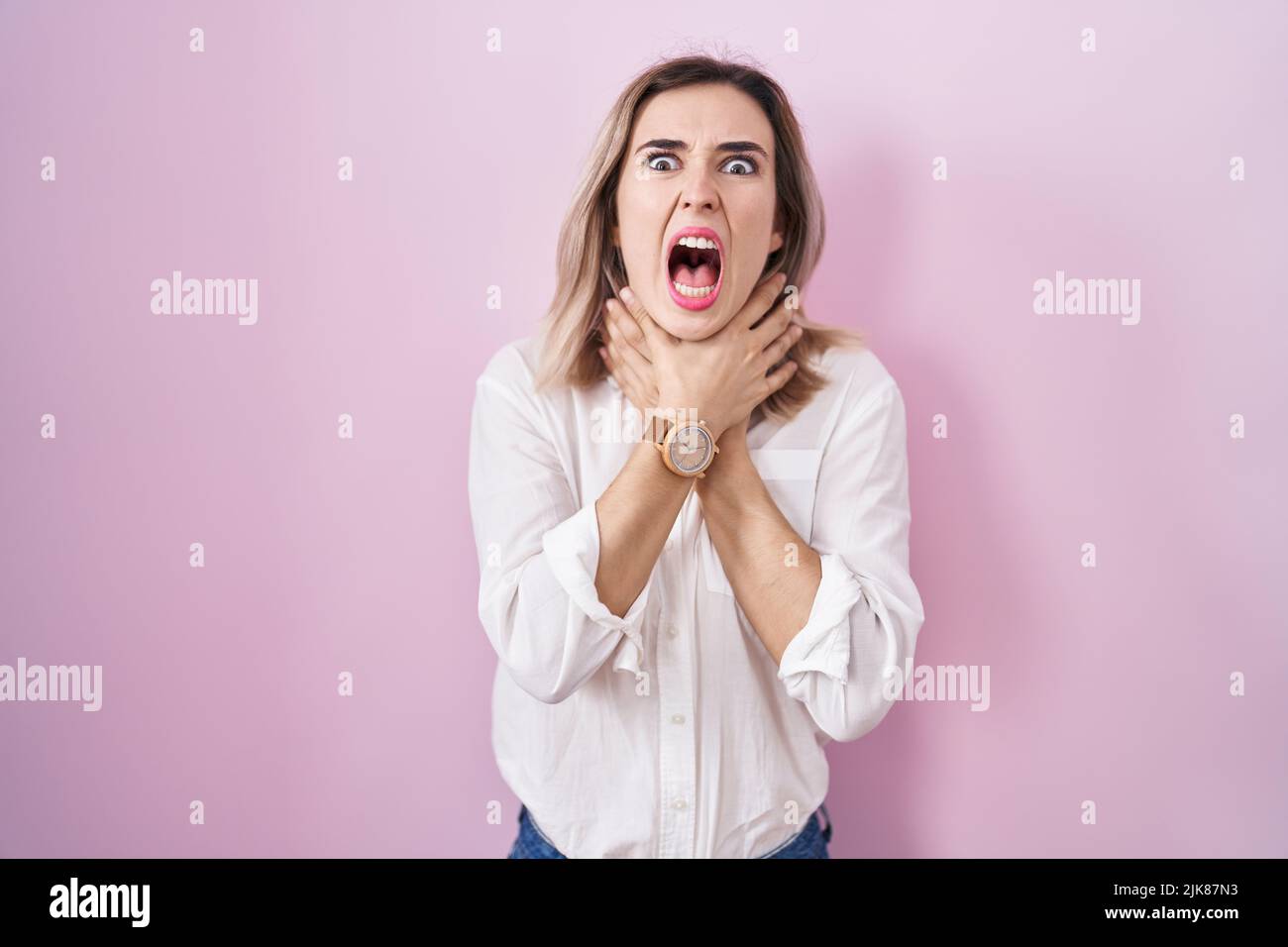 Young beautiful woman standing over pink background shouting suffocate ...