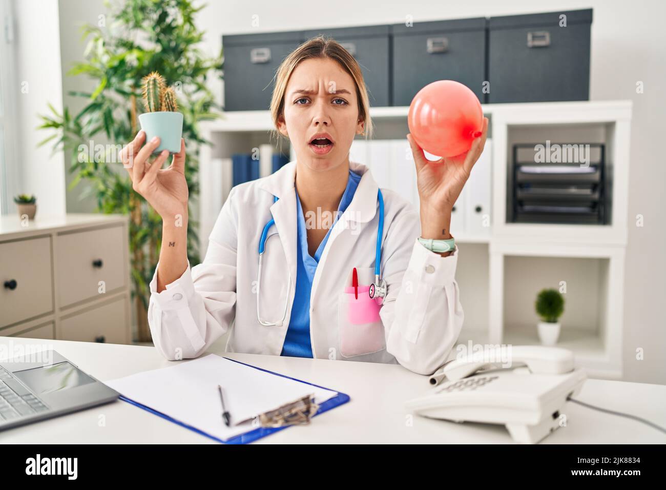 Young blonde woman wearing doctor uniform holding balloon and cactus in ...