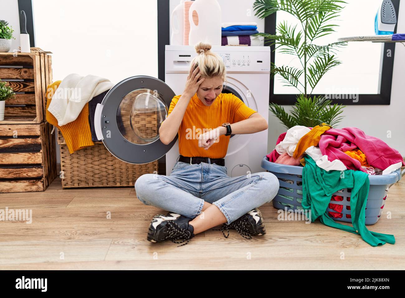 Young blonde woman doing laundry sitting by washing machine looking at ...