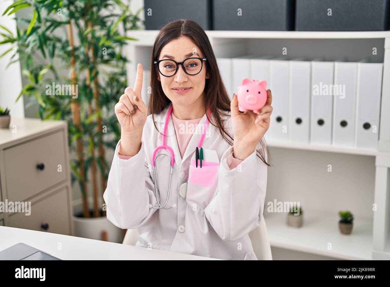 Young doctor woman holding piggy bank smiling with an idea or question ...