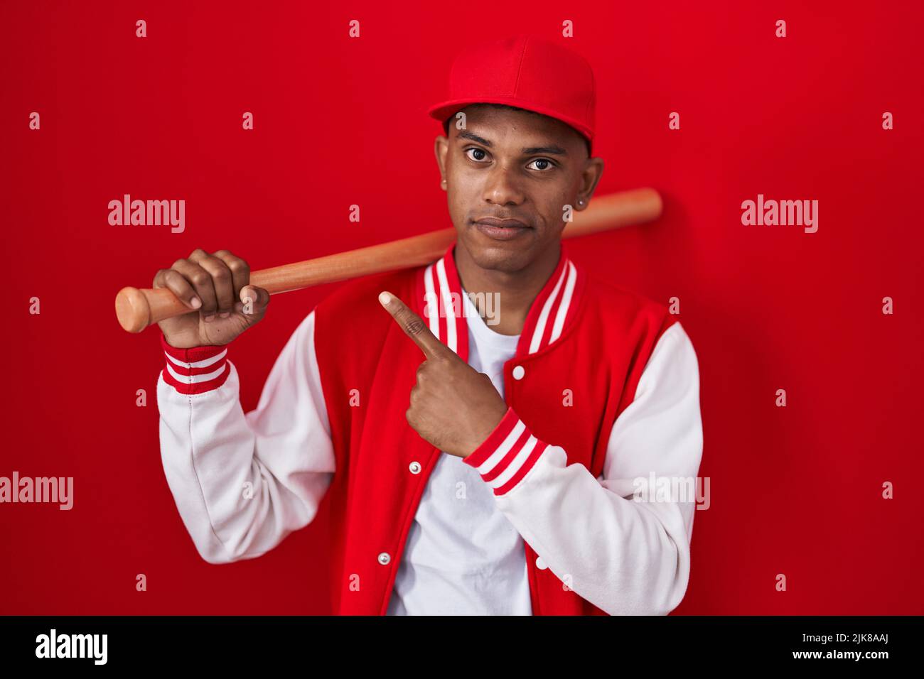 Young hispanic man playing baseball holding bat pointing with hand ...