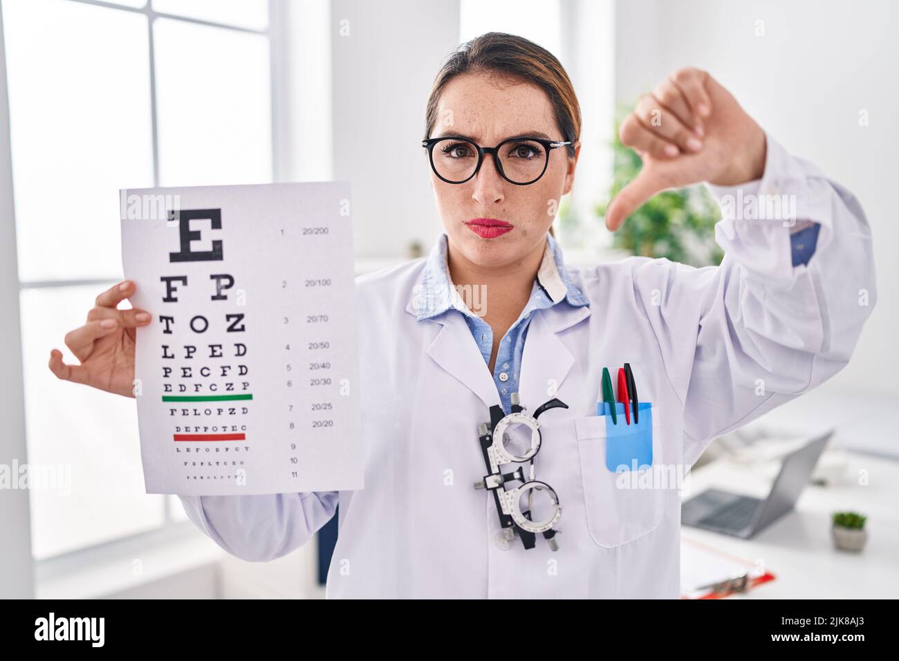 Young hispanic optician woman holding medical exam with angry face ...