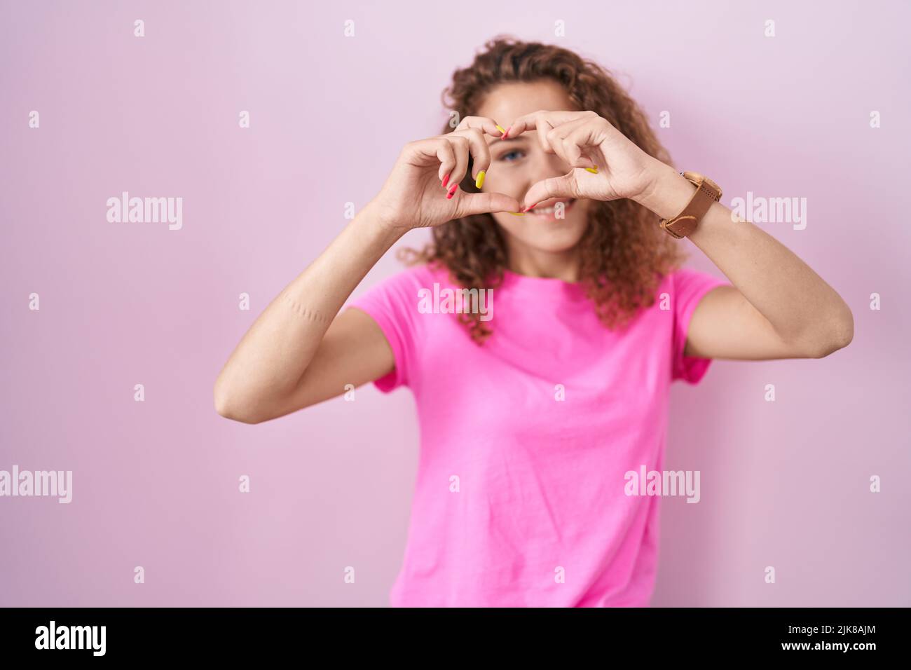 Young caucasian woman standing over pink background doing heart shape ...