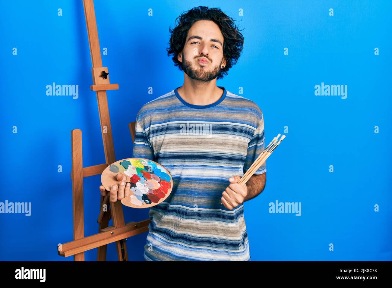 Handsome hispanic man standing drawing with palette by painter easel ...